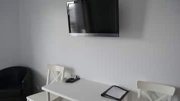 Quarto com televisão de tela plana na parede, escrivaninha branca com cadeira, controle remoto e tablet.