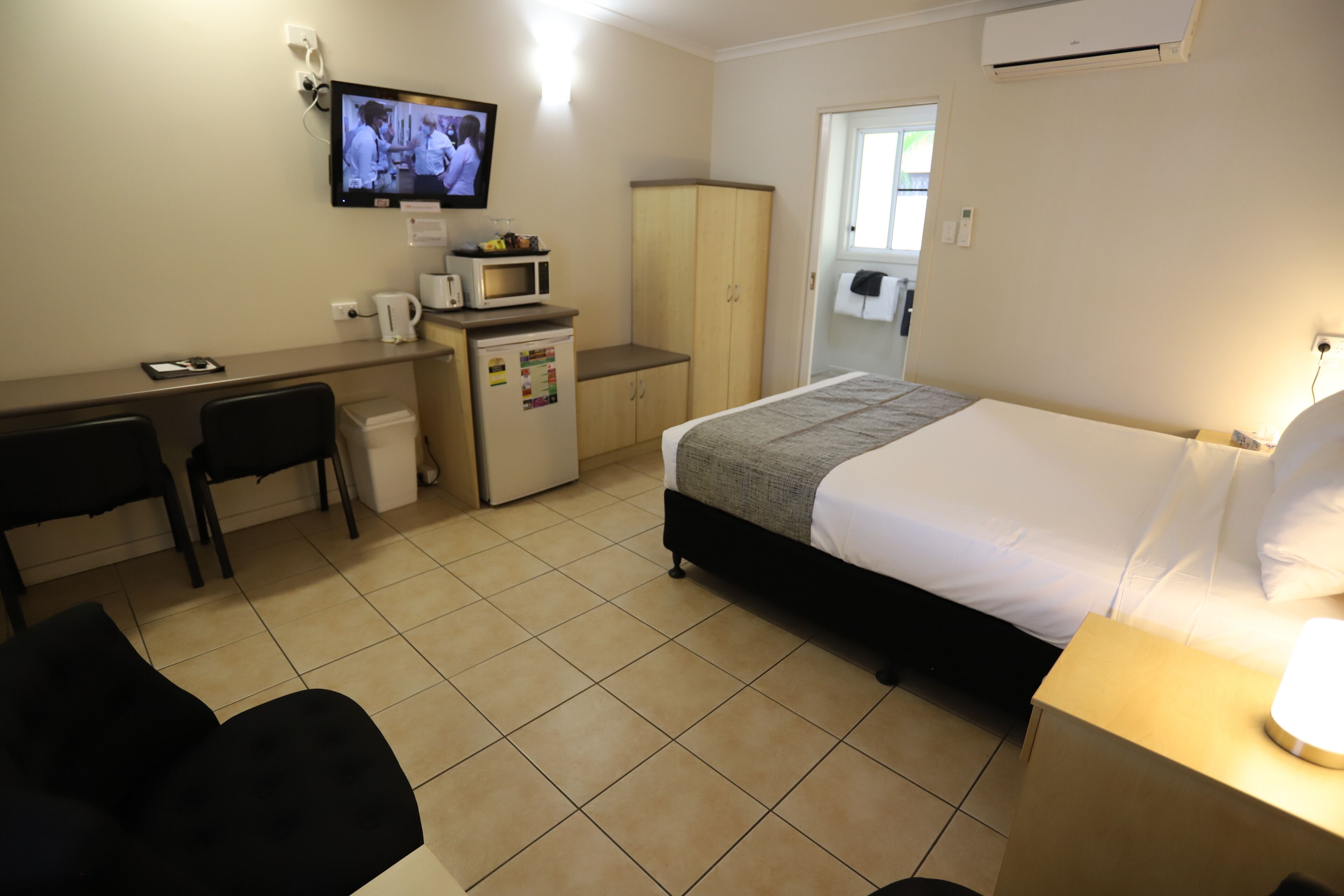 Quarto de hotel com cama, escrivaninha, cadeiras, televisão e cozinha compacta.