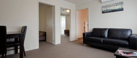 Uma sala de estar com um sofá de couro preto, uma mesa de jantar com cadeiras e uma mesa de centro. Há uma porta que dá para outro quarto com uma cama.