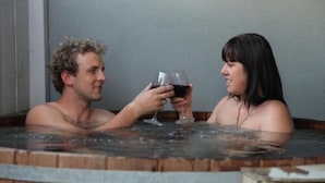 Duas pessoas em uma banheira de hidromassagem com borda de madeira, segurando copos de vinho.