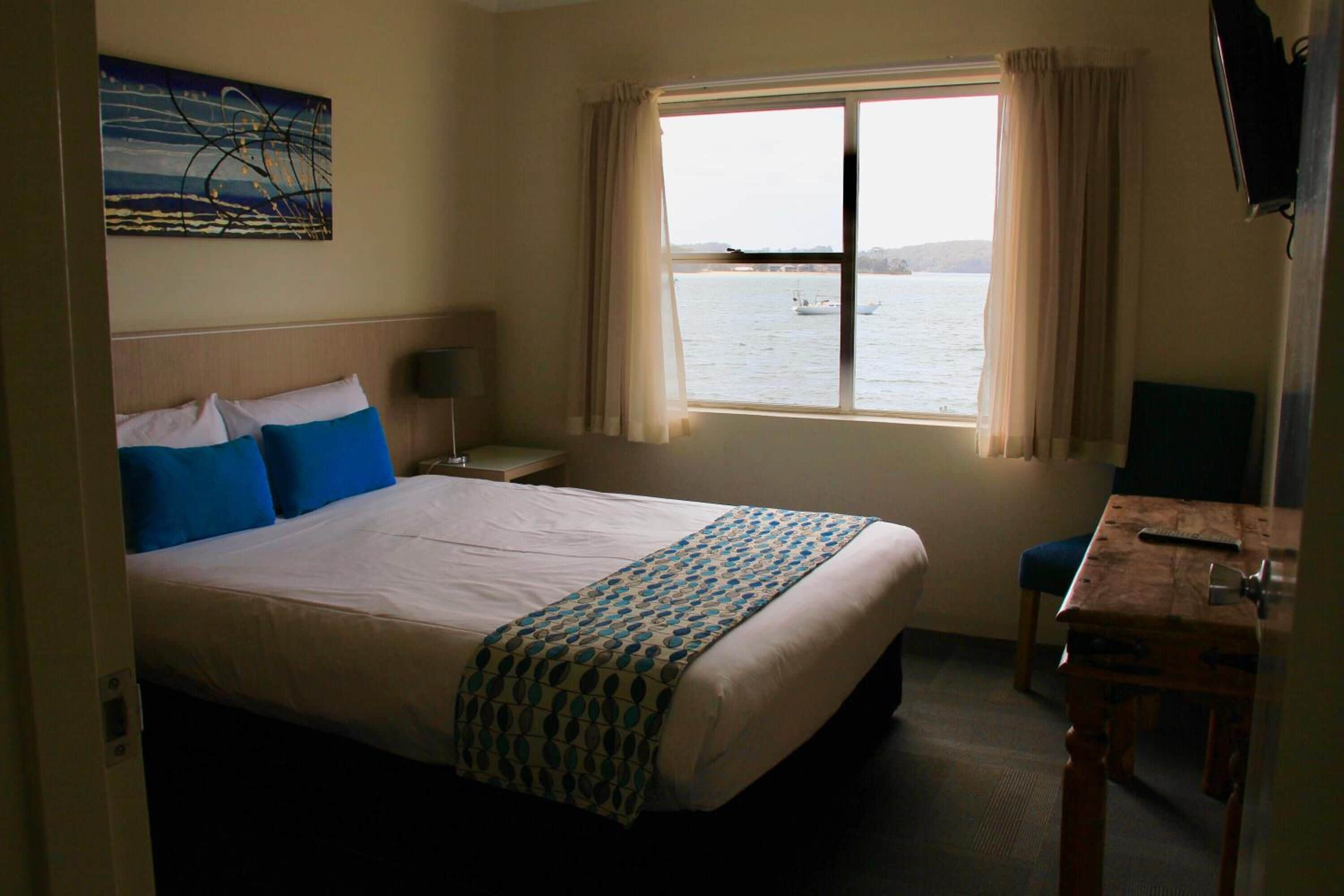 Quarto de hotel com cama, escrivaninha, cadeira, janela com vista para a água e um quadro na parede.