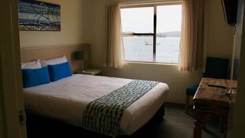 Quarto de hotel com cama, escrivaninha, cadeira, janela com vista para a água e um quadro na parede.