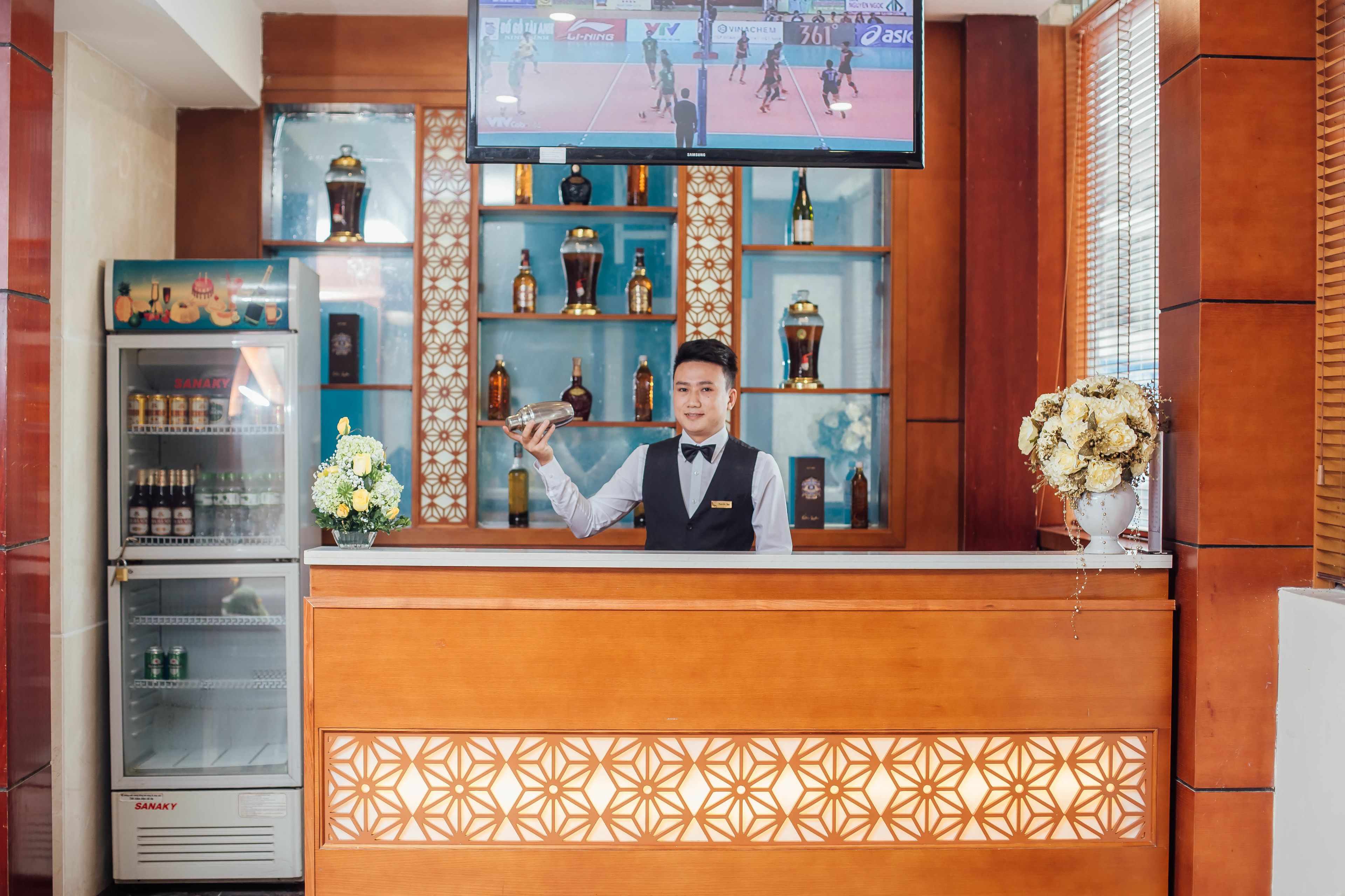 Uma área de recepção de hotel com um balcão de madeira, uma TV montada acima e prateleiras com várias garrafas e uma geladeira.