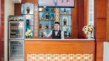 Uma área de recepção de hotel com um balcão de madeira, uma TV montada acima e prateleiras com várias garrafas e uma geladeira.