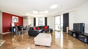 Uma sala de estar moderna com um sofá de couro preto, um banquinho branco, uma área de jantar com mesa e cadeiras, e uma grande TV de tela plana.