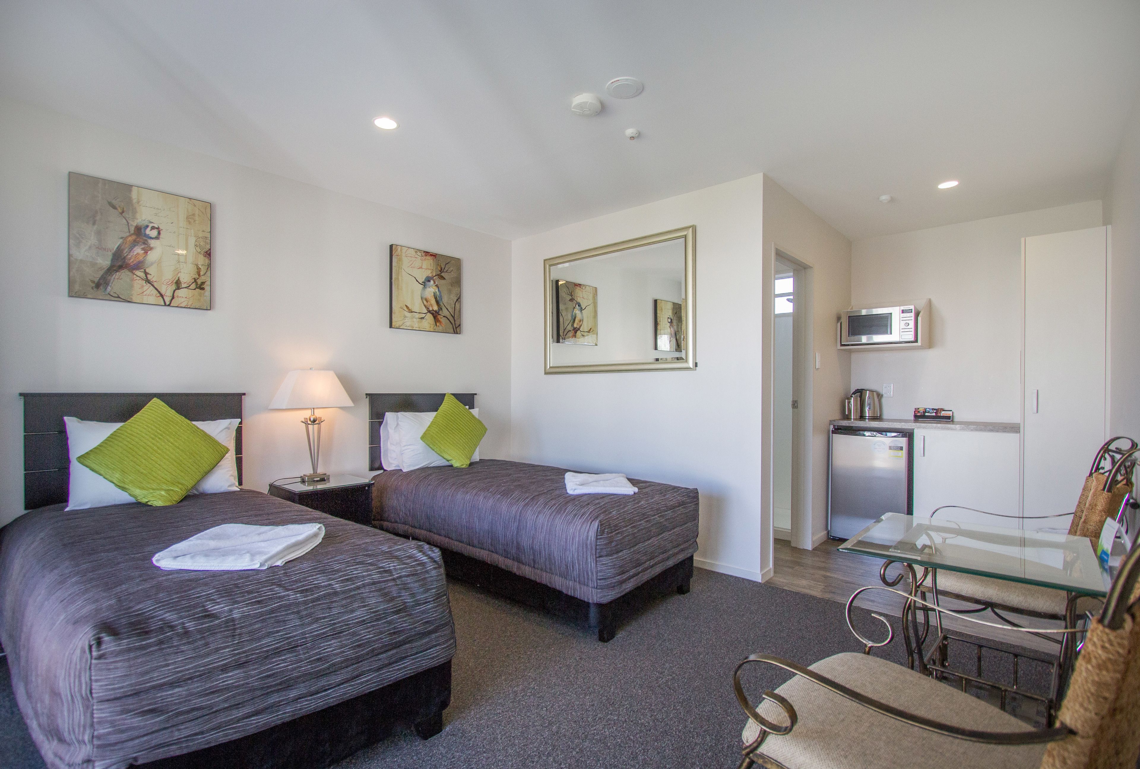 Quarto de hotel com duas camas, área de estar com mesa de vidro, micro-ondas e pequena cozinha.