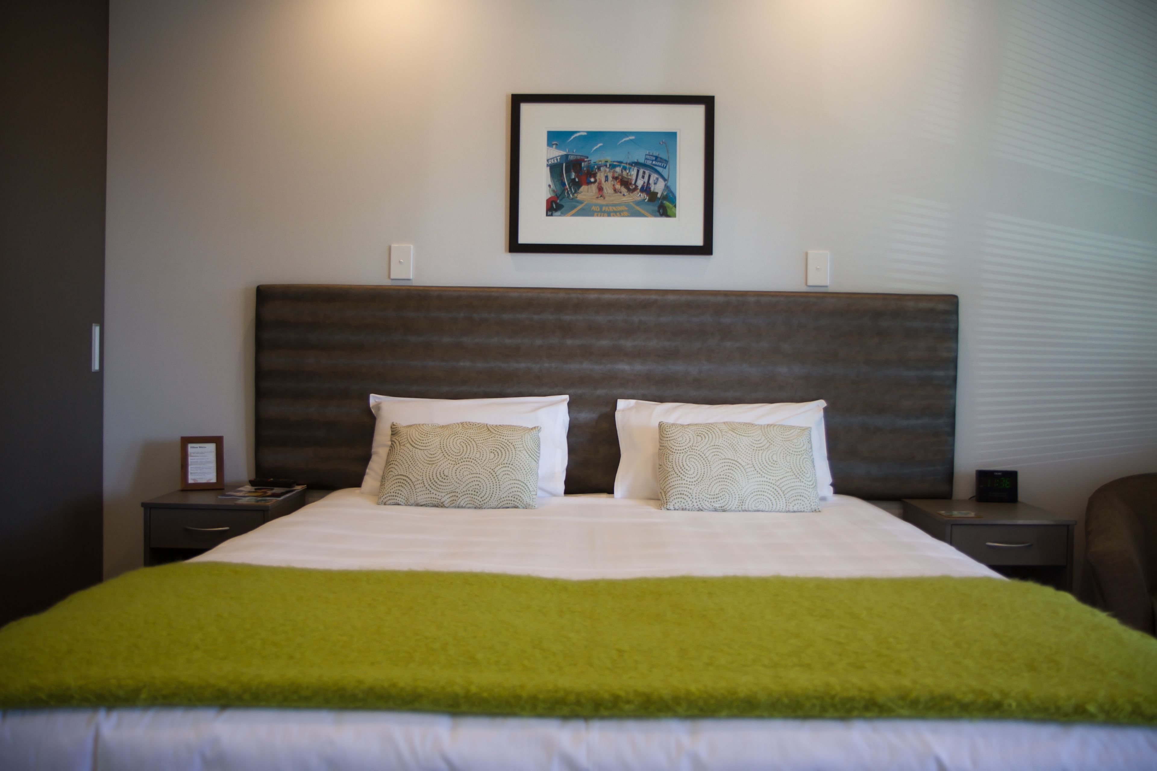 Quarto de hotel com uma cama grande, duas almofadas, um cobertor verde, um quadro na parede e uma mesa de cabeceira com abajur.