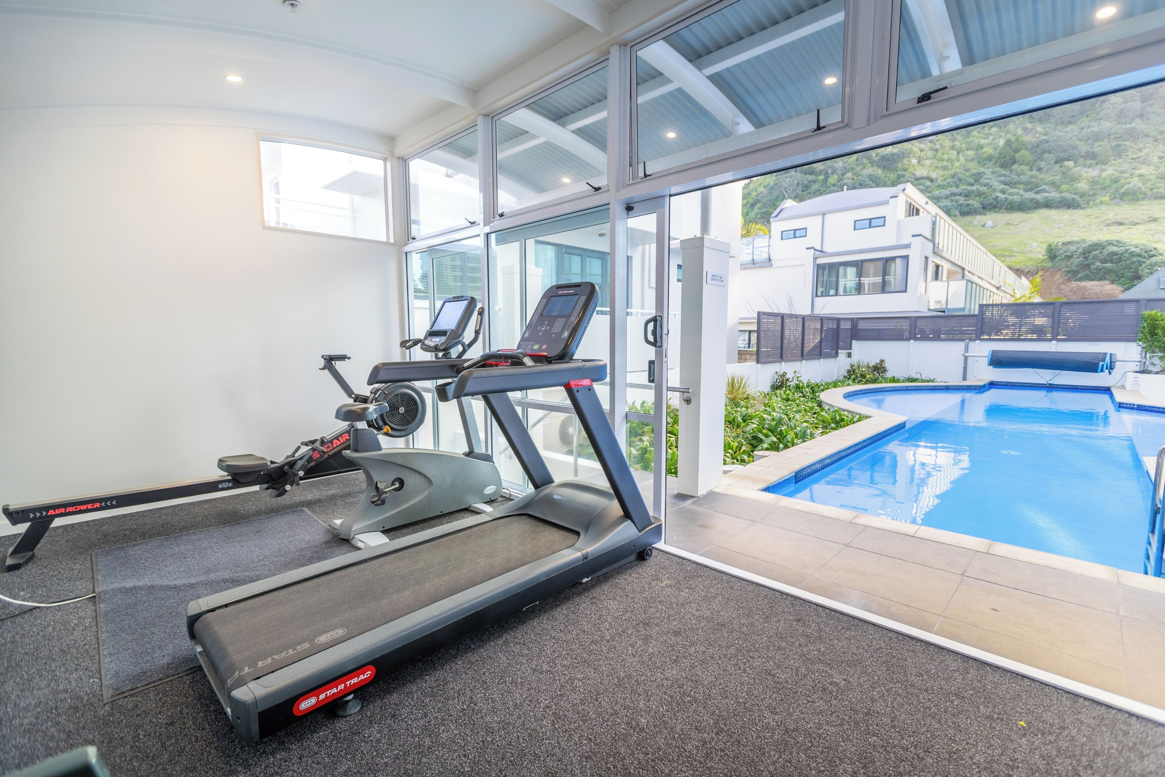 Ginásio com esteiras e bicicletas ergométricas, piscina e vista para uma casa moderna.
