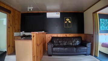 Quarto com parede de painel de madeira, sofá de couro preto, pequena cozinha com fogão e ar-condicionado instalado no teto.
