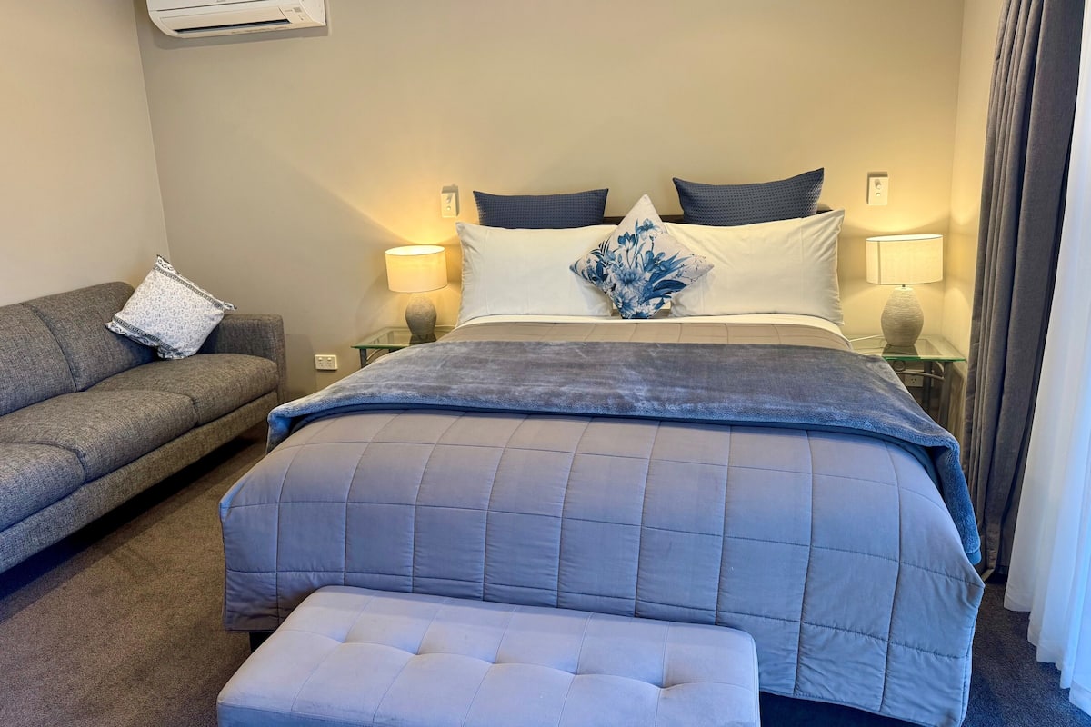 Quarto de hotel com cama, sofá, duas luminárias de cabeceira, ar condicionado e janela com cortinas.