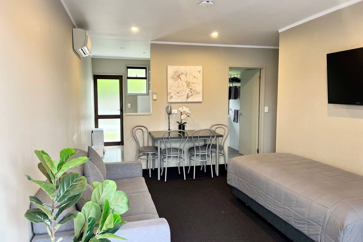 Quarto compacto com sofá cinza, uma pequena mesa de jantar com cadeiras, uma cama de solteiro e televisão na parede.