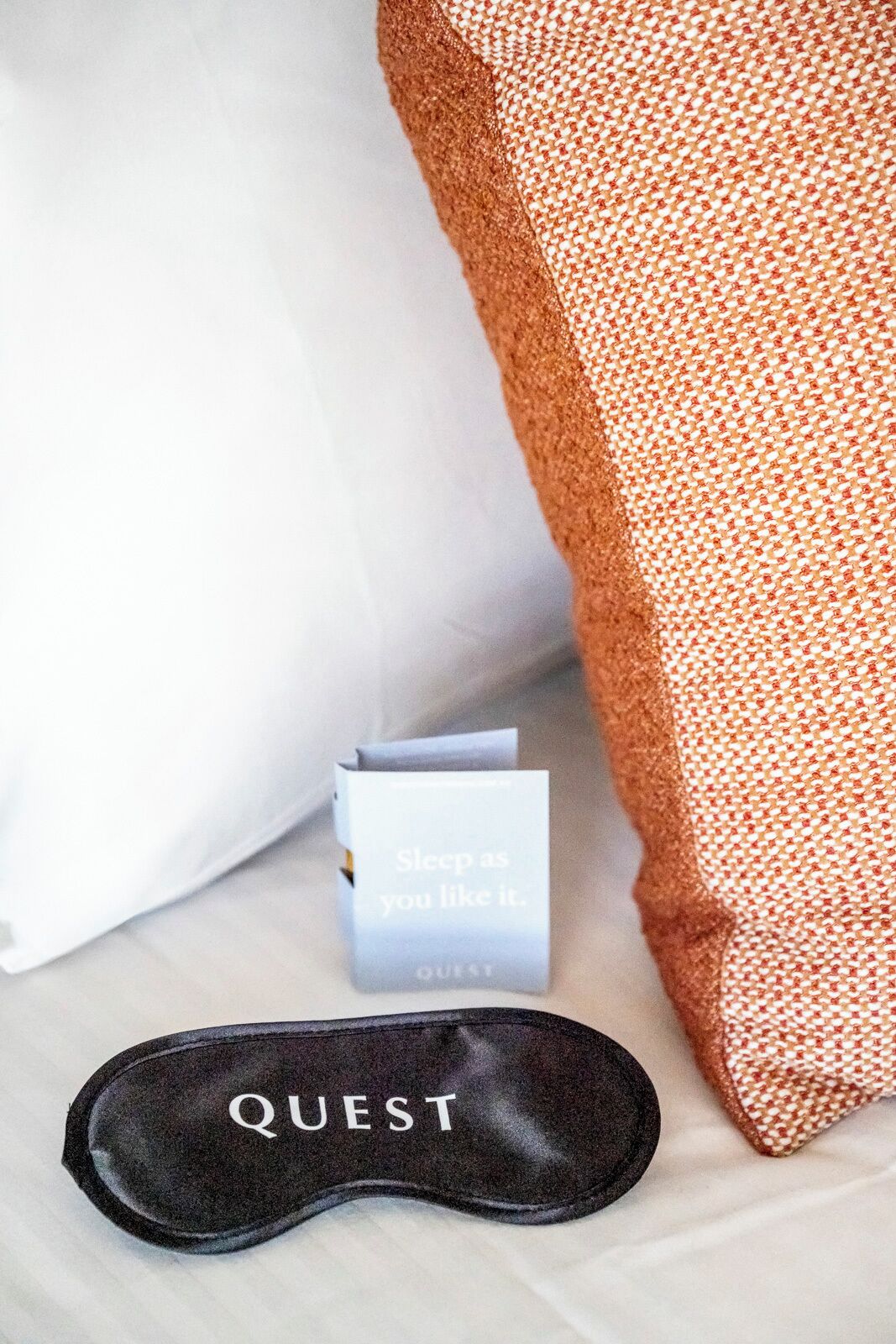 Uma cama com travesseiro branco, travesseiro castanho e máscara para os olhos preta com a inscrição 'QUEST', além de um pequeno cartão com o texto 'Durma como quiser'.