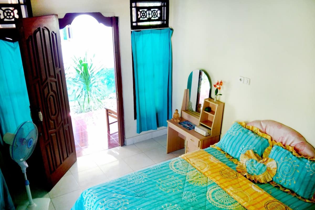 Quarto com cama, escrivaninha, cadeira, ventoinha e vista para o exterior.