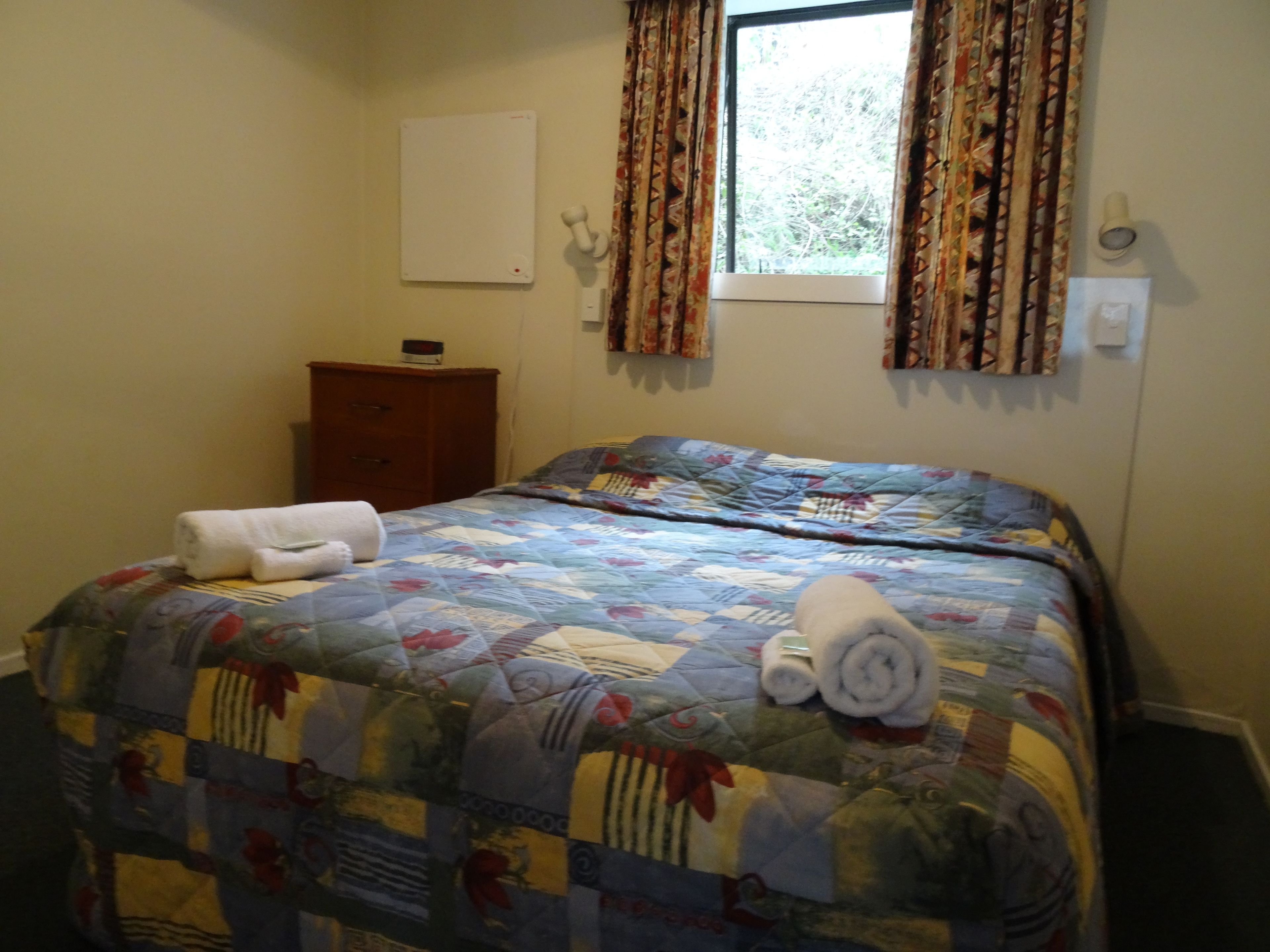 Quarto com cama, mesa de cabeceira, janela com cortinas e luminária na parede.