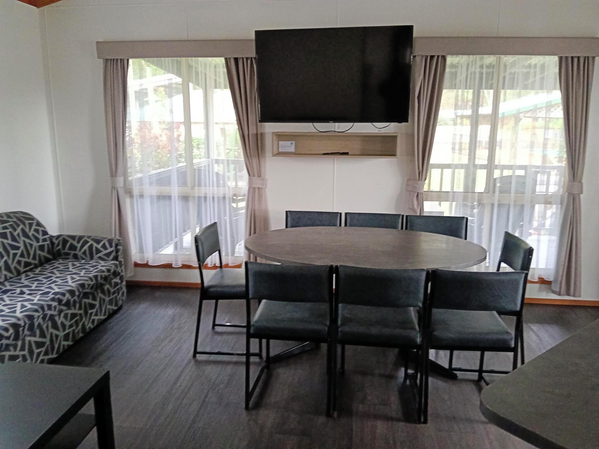 Um quarto com uma mesa redonda cercada por cadeiras, um sofá e uma televisão fixada na parede.
