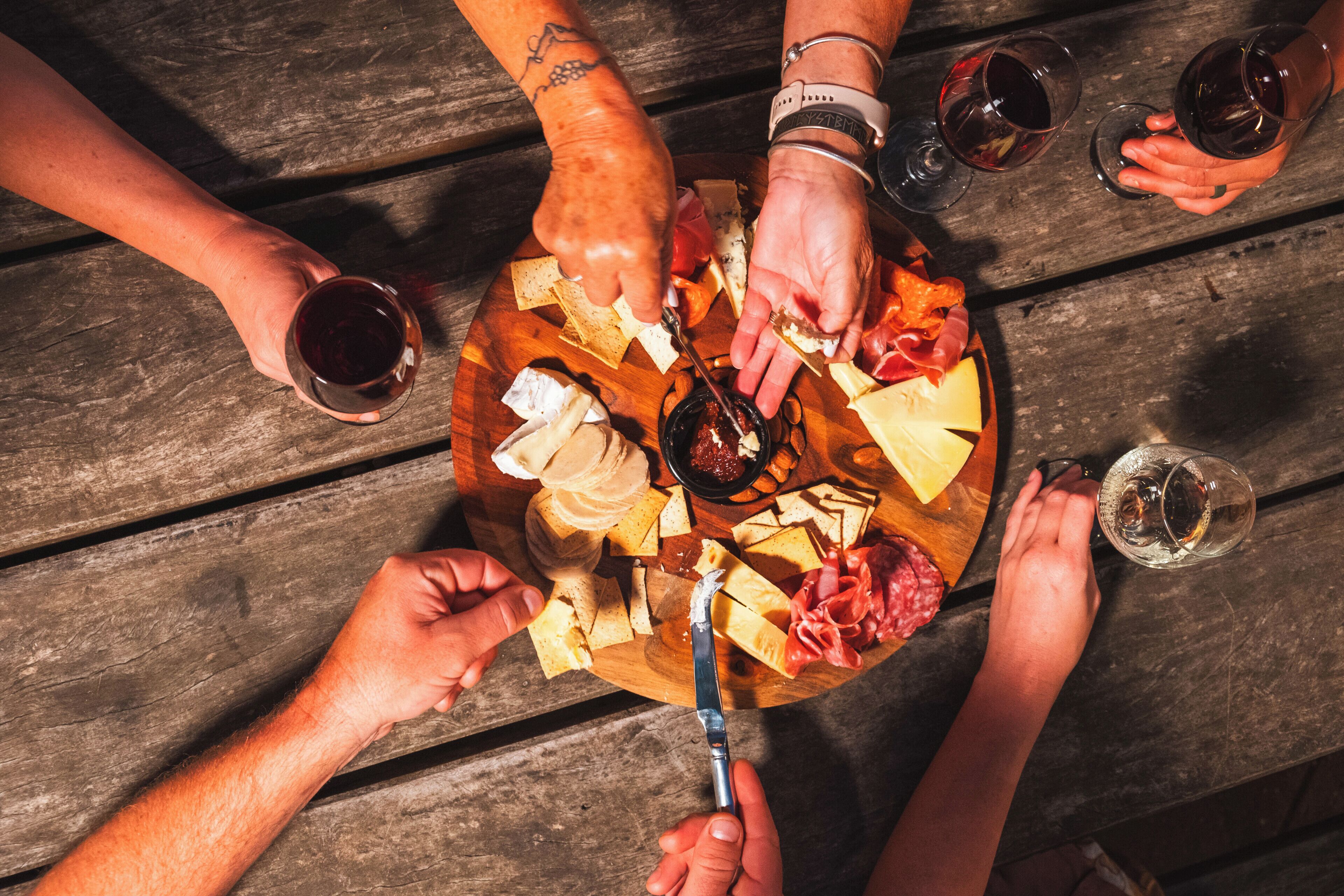 Um grupo de pessoas reunido em torno de uma mesa de madeira com diversos queijos, carnes e biscoitos.