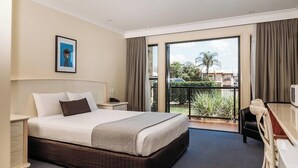 Quarto de hotel com uma cama grande, uma escrivaninha, uma cadeira e varanda com vista para palmeiras e edifícios.