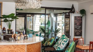 Interior de restaurante com área de bar, cadeiras de madeira com almofadas tropicais e um menu em lousa listando preços.