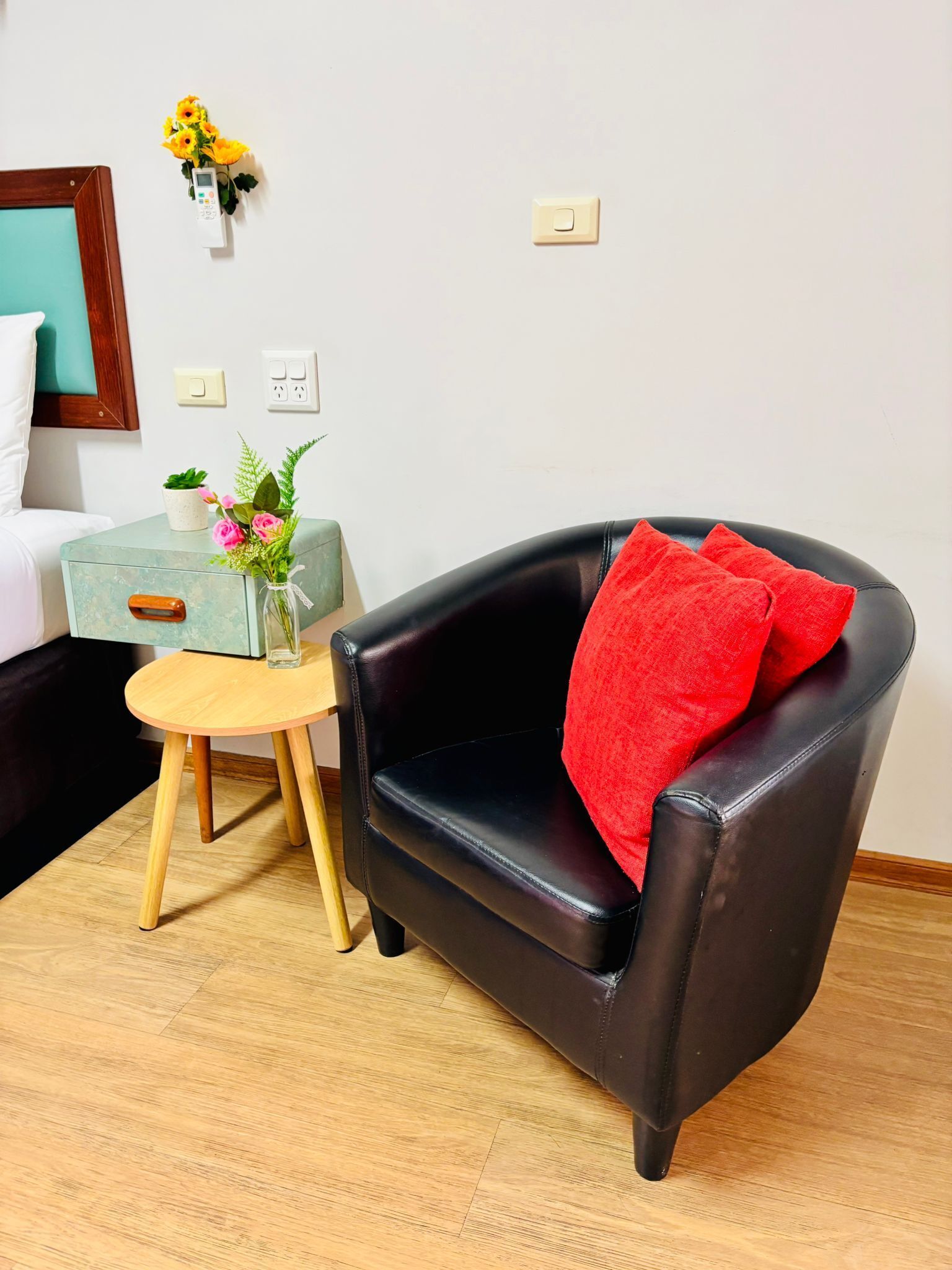 Quarto com sofá de couro preto, uma pequena mesa de madeira, um criado-mudo verde-esmeralda com uma planta e uma cama com roupa de cama branca.