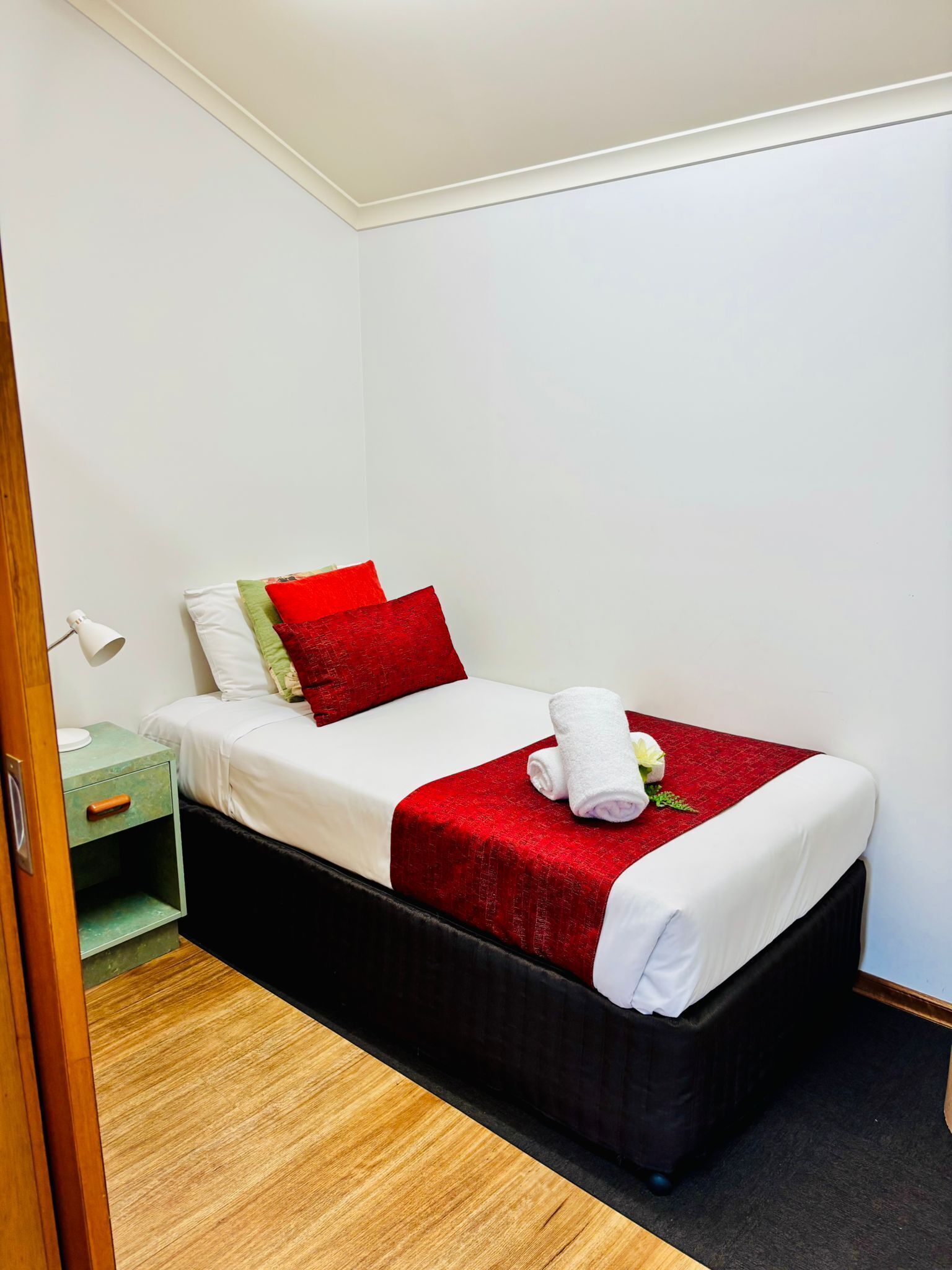 Cama bem arrumada com manta e travesseiros vermelhos, uma toalha branca e uma pequena mesa de cabeceira com abajur.