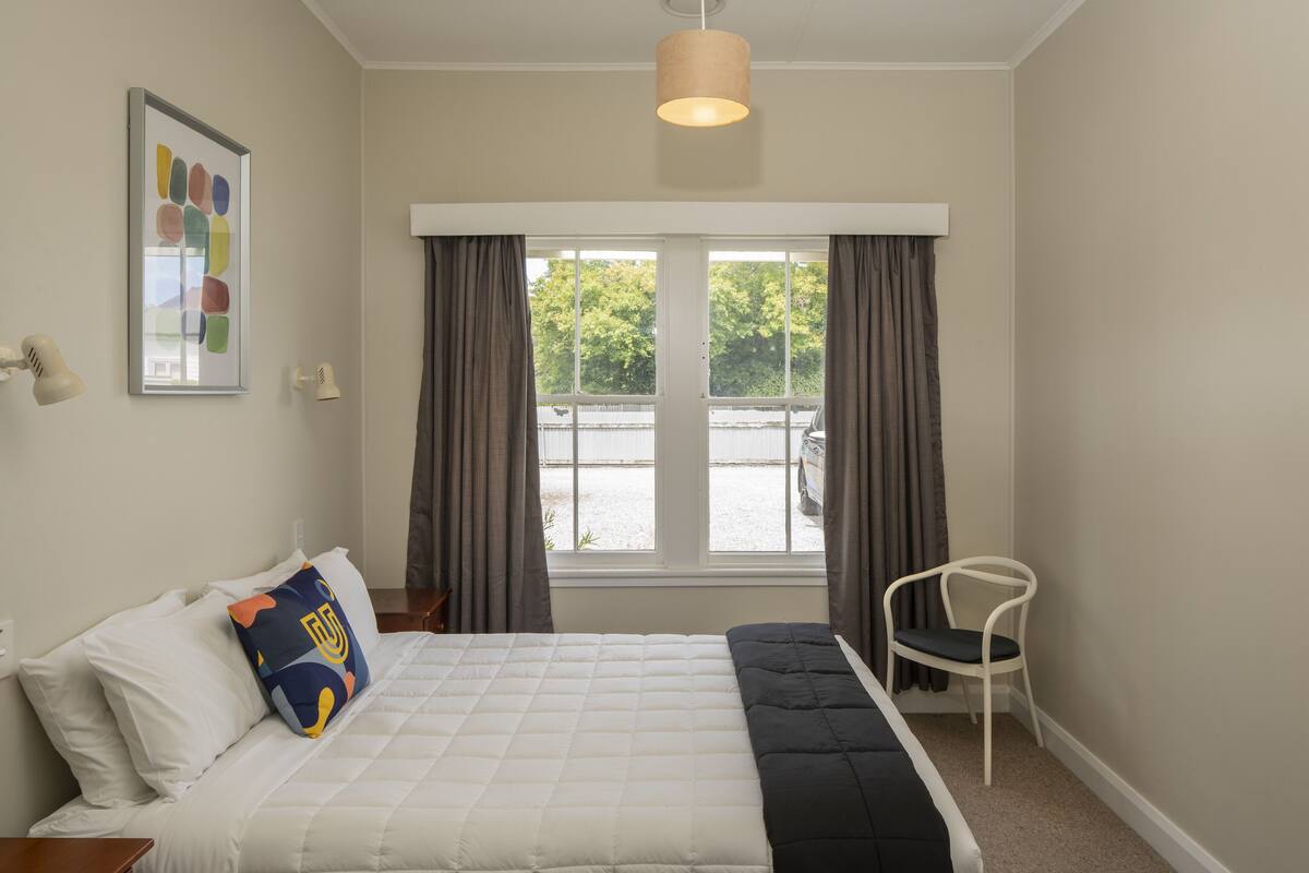 Quarto com cama, uma cadeira, janela com cortinas e luminária pendente.