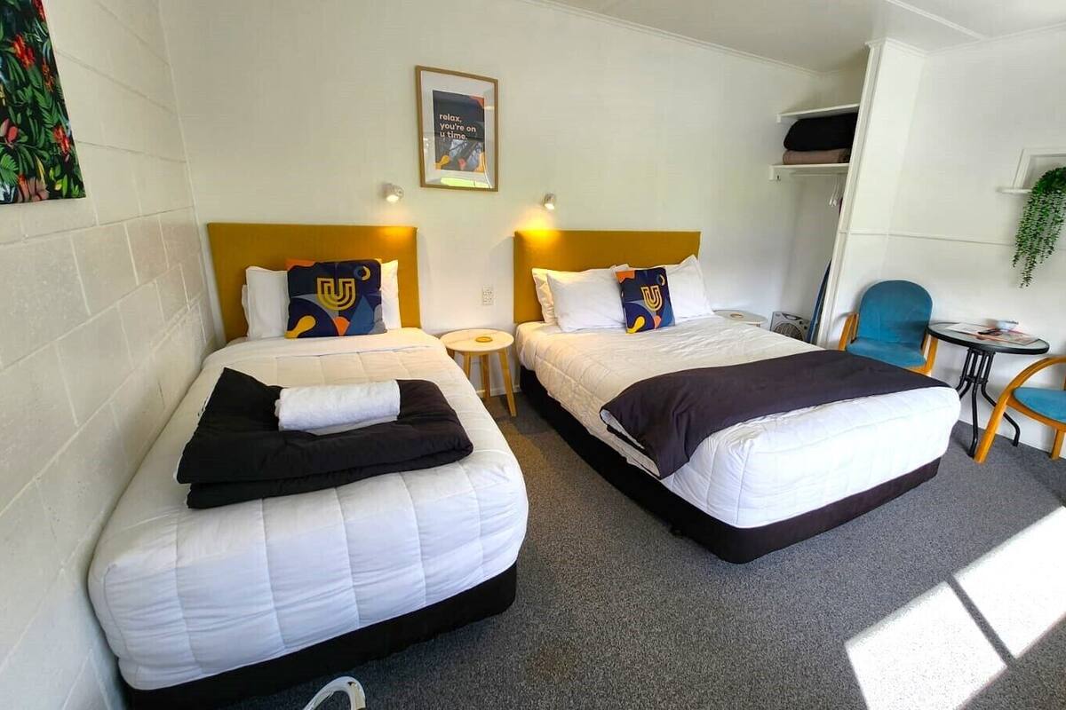 Quarto de hotel com duas camas, uma mesinha, uma cadeira e um prateleira fixada na parede com uma planta.