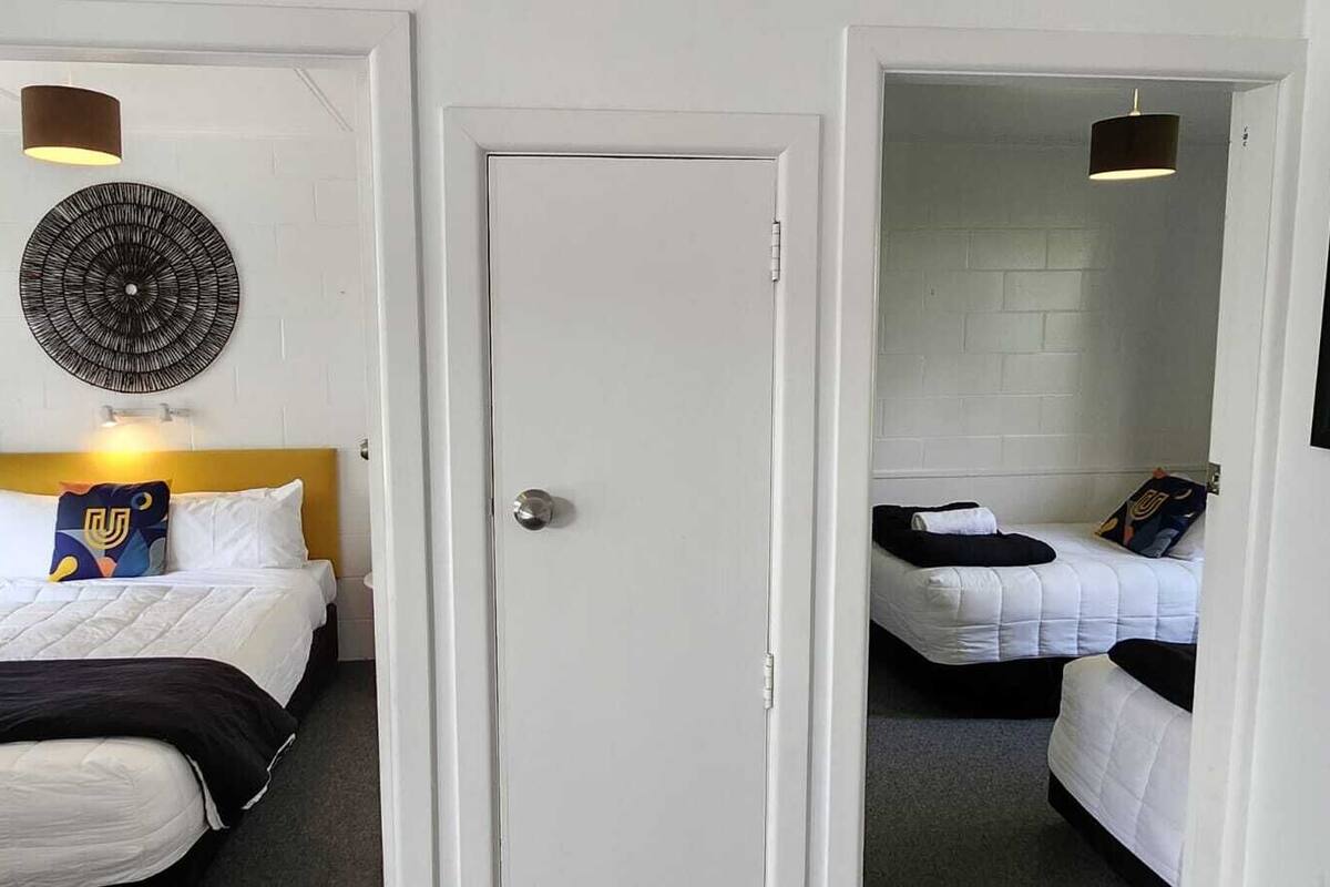 Quarto de hotel com cama, mesa de cabeceira, luminária na parede e porta para outro cômodo.