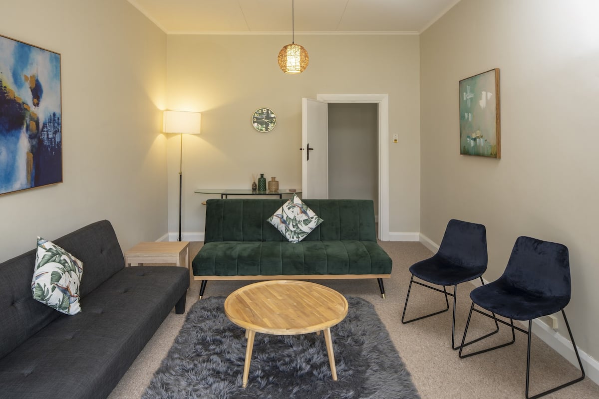 Uma sala de estar moderna com um sofá verde escuro, duas poltronas azul-escuras, uma mesa de centro de madeira e um tapete macio. Uma luminária, um relógio e um quadro estão nas paredes.