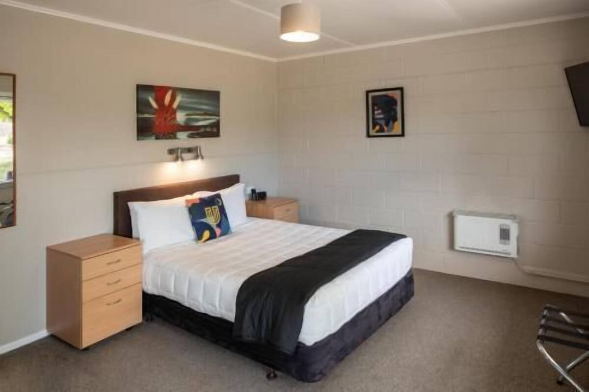 Quarto com cama, mesa de cabeceira, cômoda, aquecedor fixado na parede e duas pinturas.