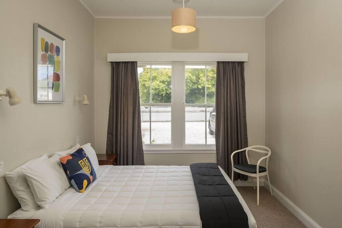 Quarto com cama, cadeira, janela com cortinas, luminária na parede e luz no teto.