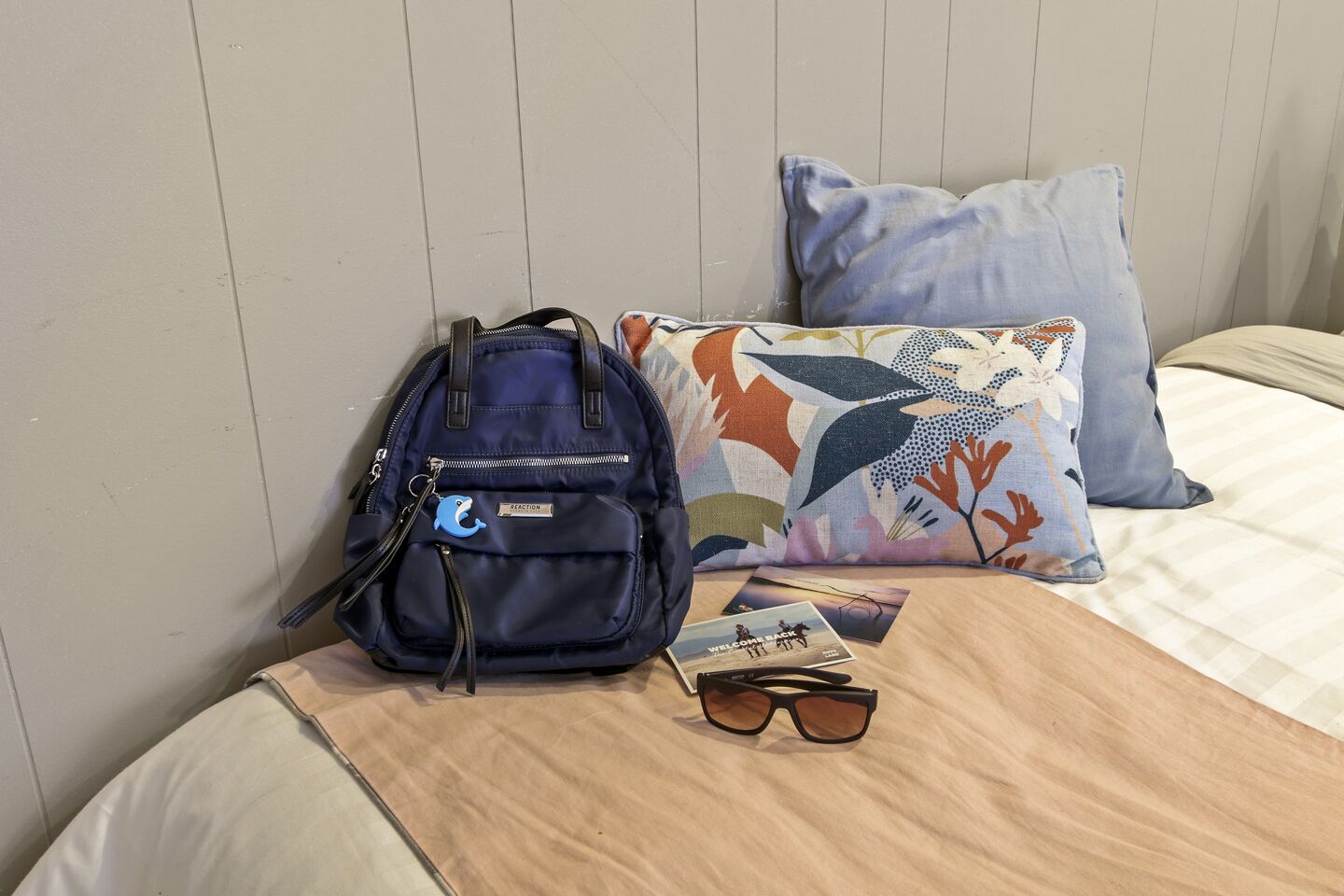 Uma cama com um travesseiro azul, uma mochila azul e um par de óculos de sol sobre uma mesa de cabeceira de madeira.