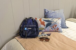 Uma cama com um travesseiro azul, uma mochila azul e um par de óculos de sol sobre uma mesa de cabeceira de madeira.