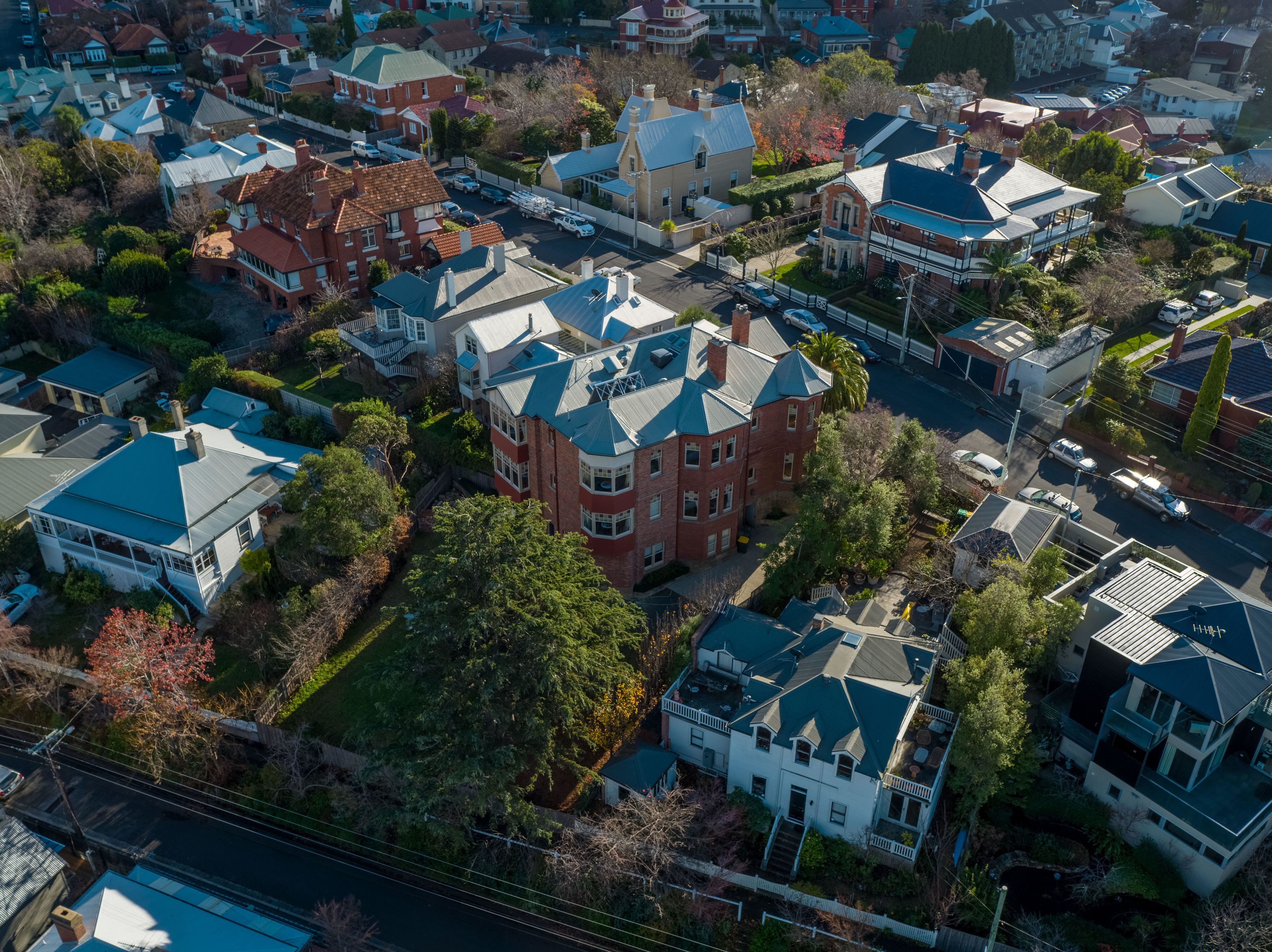 Vista aérea de uma área residencial com diversas casas, incluindo uma casa de tijolos vermelhos proeminente com uma torre e uma casa branca com telhado azul.