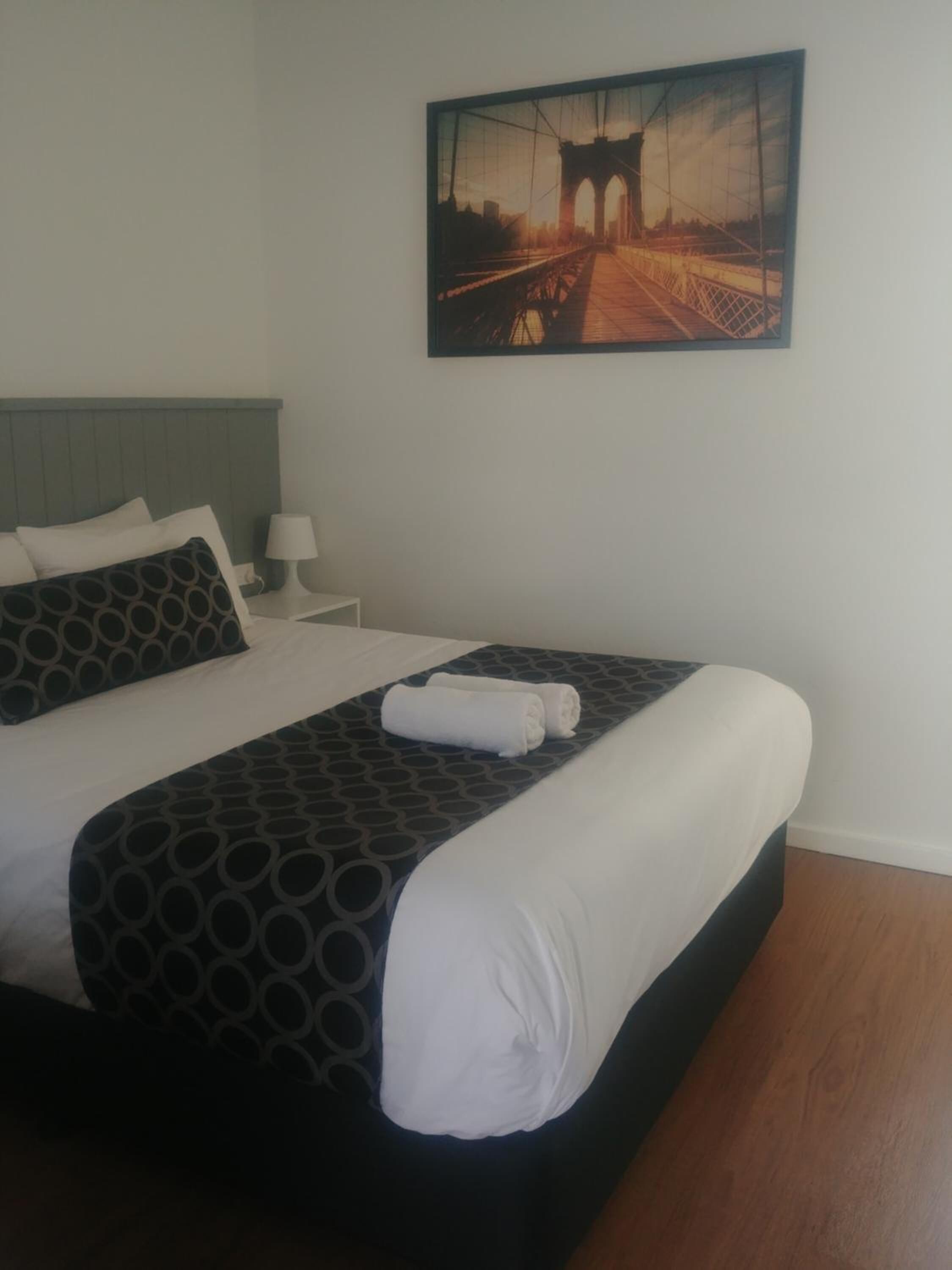 Quarto de hotel com uma cama grande, uma cabeceira, uma mesa de cabeceira com abajur e um quadro de uma ponte na parede.
