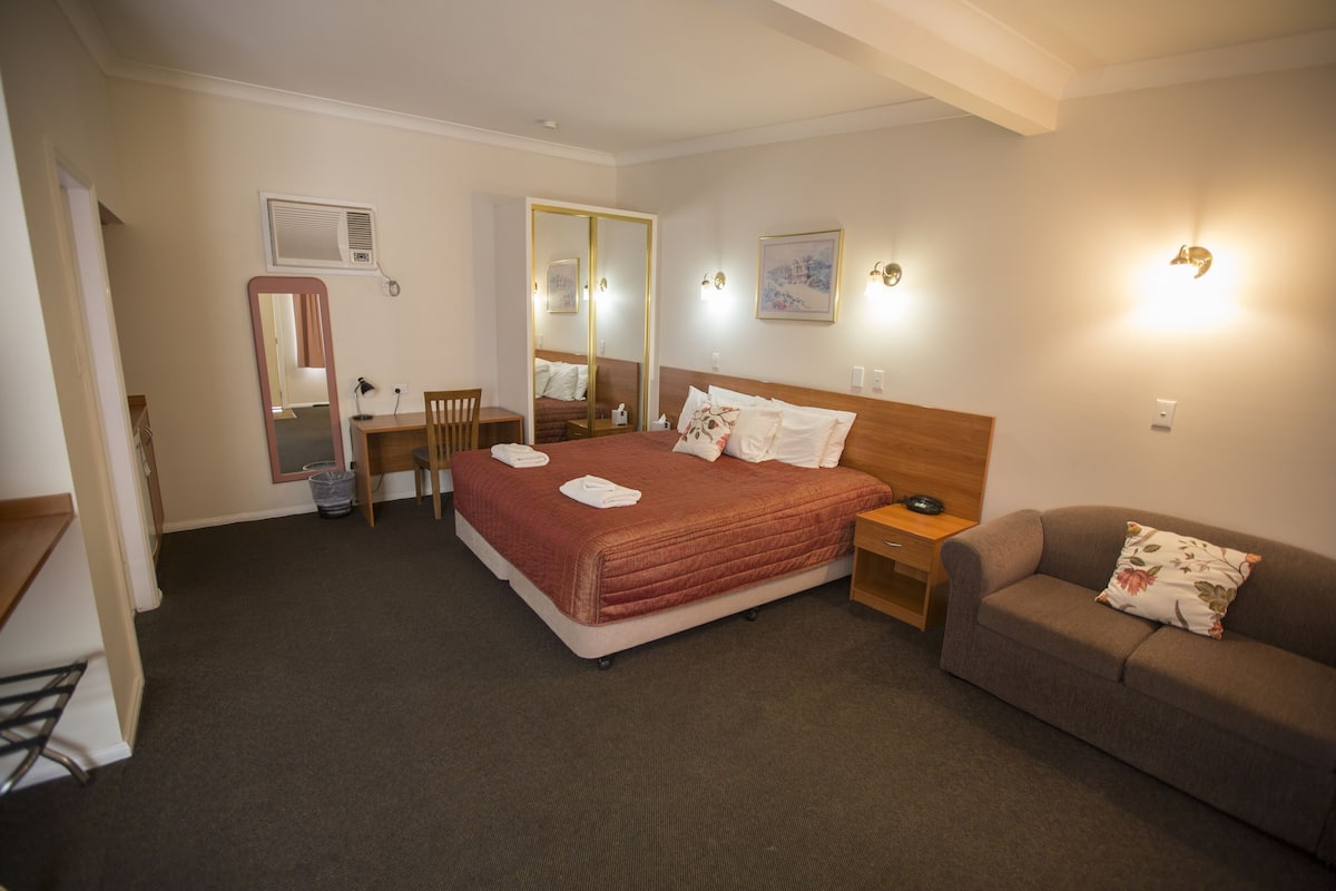 Quarto de hotel com cama, escrivaninha, cadeira, sofá e espelho.