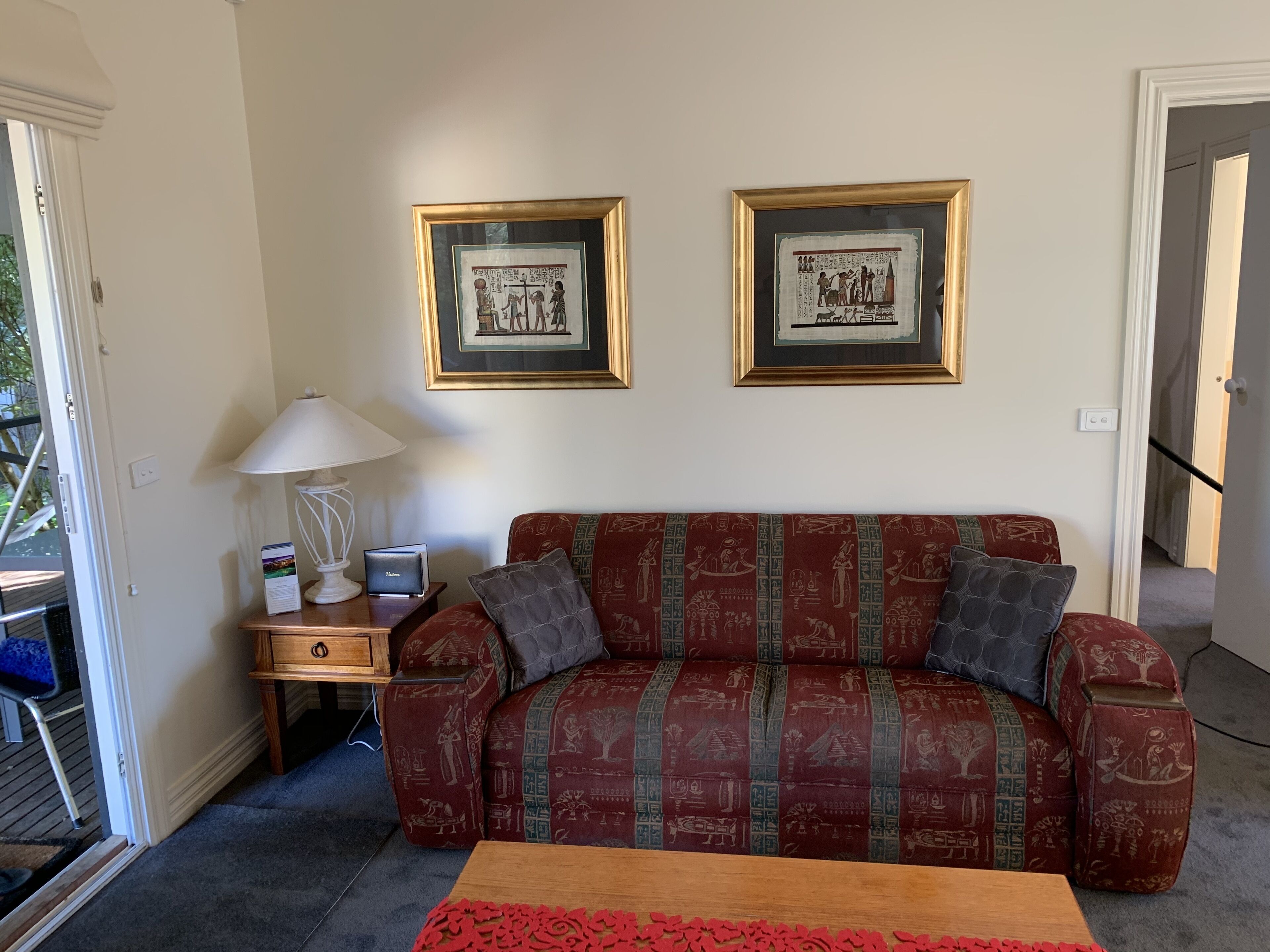 Uma sala de estar com um sofá vermelho estampado, duas imagens emolduradas na parede, uma mesa lateral de madeira com um abajur e um tapete vermelho.
