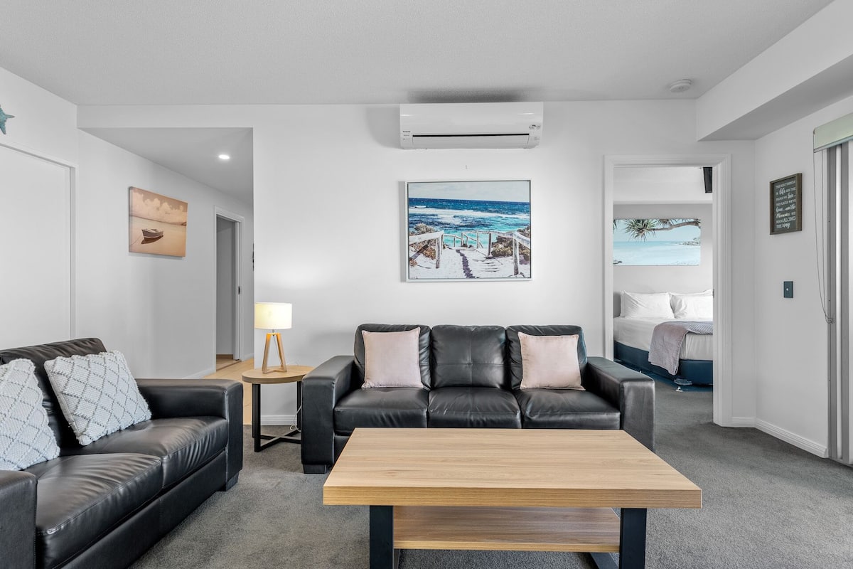 Uma sala de estar moderna com um sofá de couro preto, uma mesa de centro de madeira e uma pintura de uma cena de praia.