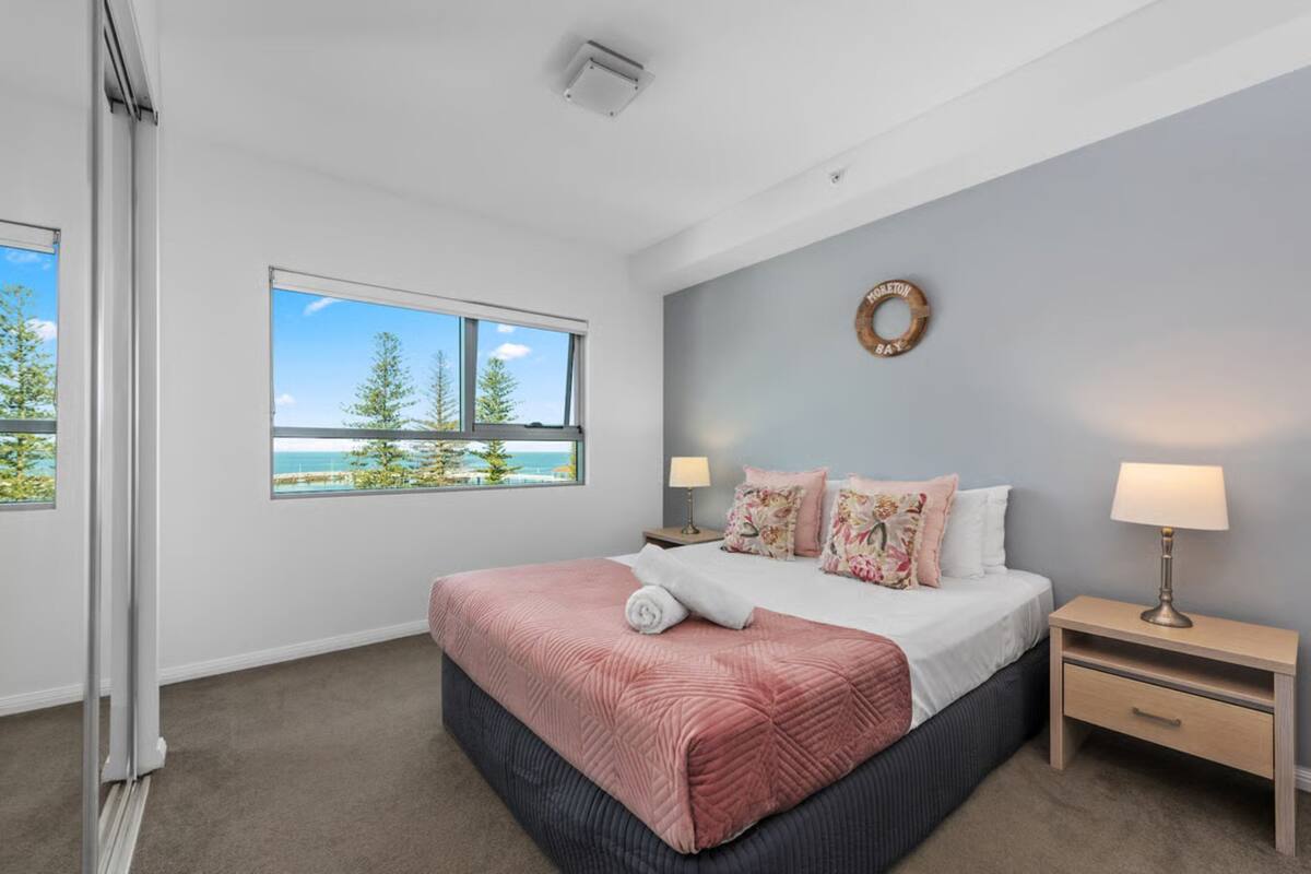 Quarto com uma cama grande, duas mesas de cabeceira, vista para o mar e uma decoração circular na parede.