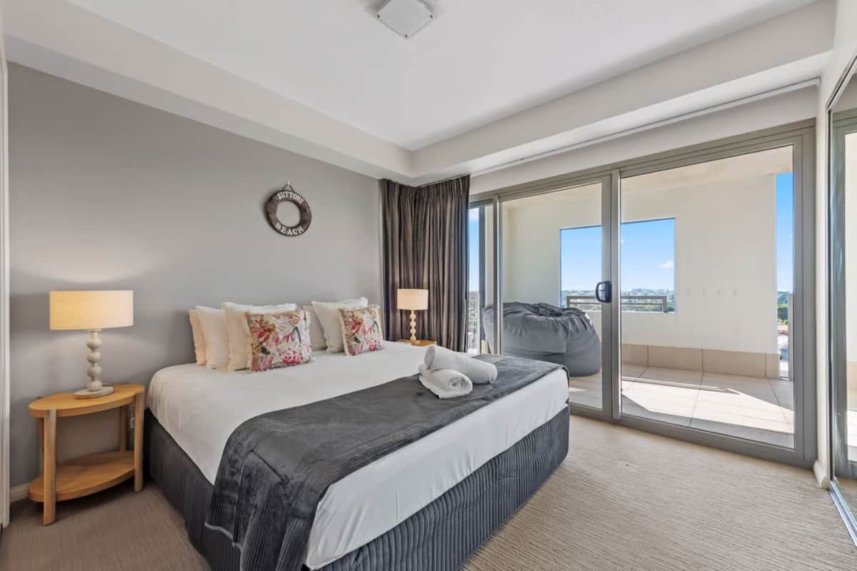 Quarto de hotel com uma cama grande, uma mesa de cabeceira, um objeto decorativo na parede e uma porta de vidro deslizante que dá para uma varanda.