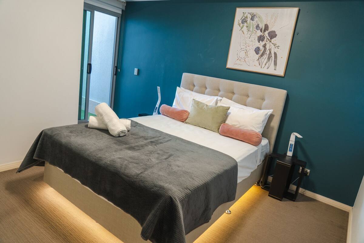 Cama bem arrumada com colcha cinza, edredom branco e várias almofadas em tons de rosa e verde. Uma obra de arte floral emoldurada está pendurada na parede verde-água acima da cama.