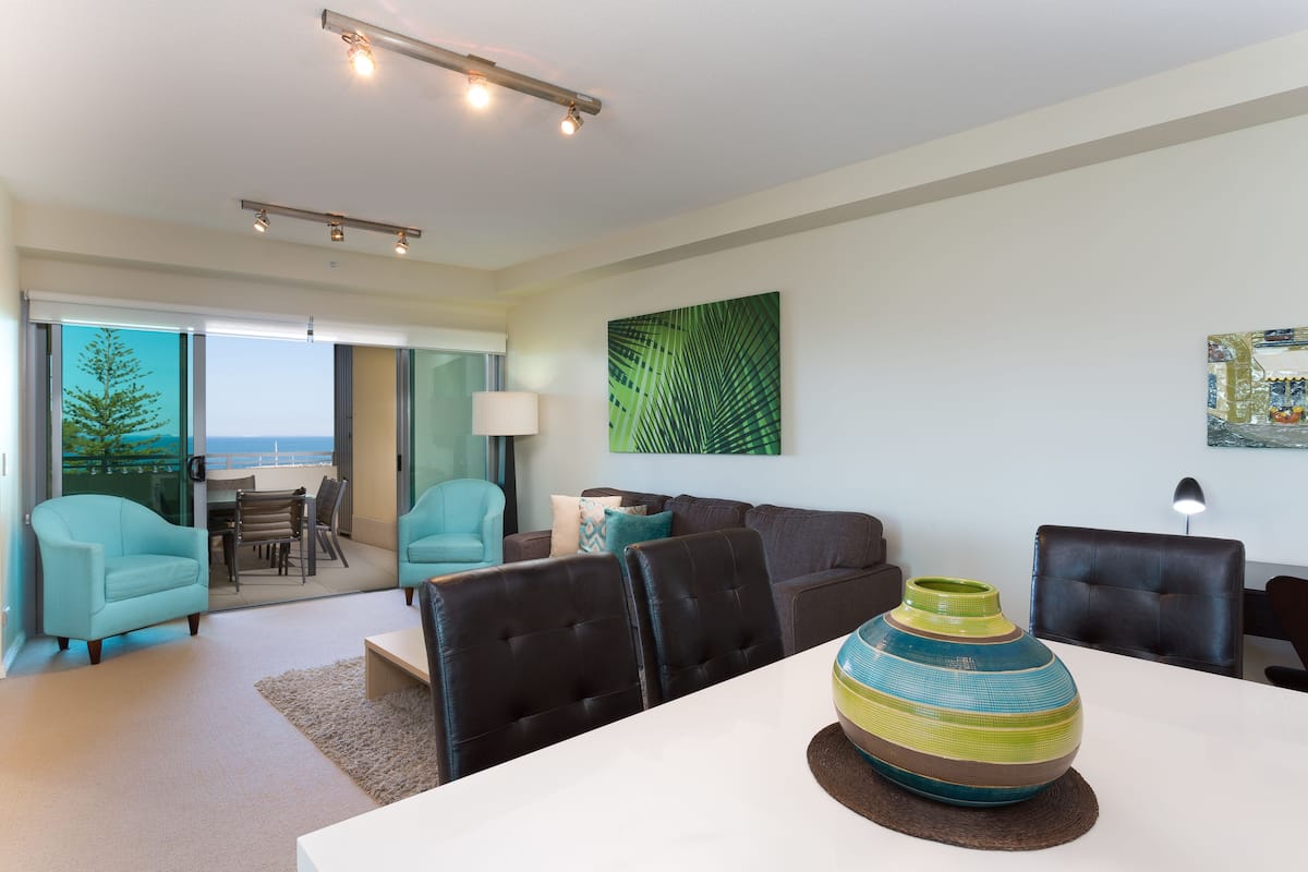 Uma sala de estar moderna com área de jantar, uma grande janela e uma obra de arte com tema tropical.