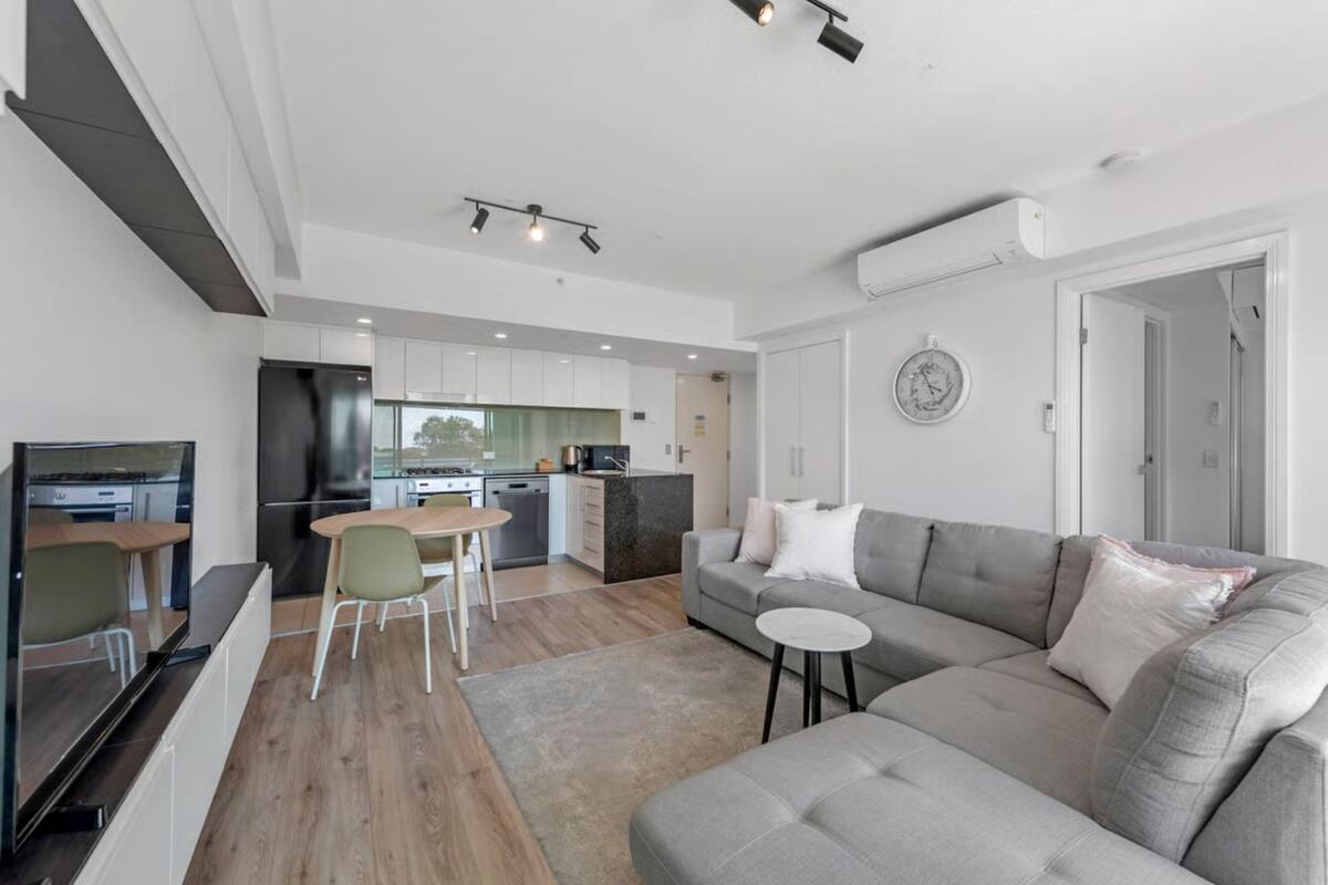 Um espaço de estar moderno com um sofá cinzento, uma área de jantar com mesa e cadeiras, e uma cozinha com armários brancos e uma geladeira de aço inoxidável.