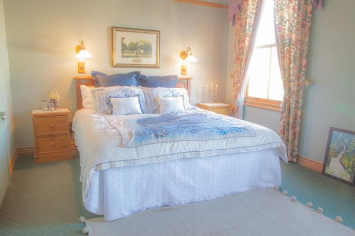 Quarto com uma cama, duas mesas de cabeceira, um criado-mudo, janela com cortinas e um quadro na parede.
