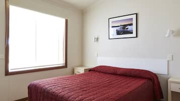 Quarto com uma janela grande, uma cama com colcha vermelha, mesa de cabeceira e um pequeno criado-mudo, além de um quadro pendurado na parede.