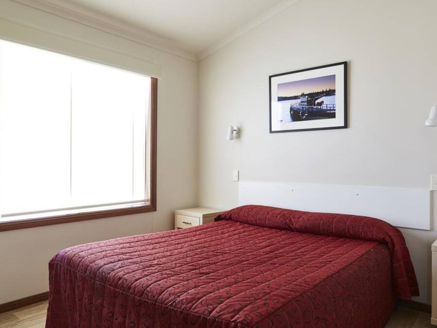 Quarto com uma janela grande, uma cama com colcha vermelha, mesa de cabeceira e um pequeno criado-mudo, além de um quadro pendurado na parede.