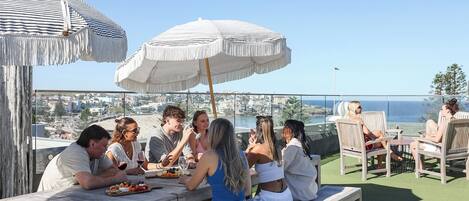 Um grupo de pessoas jantando em uma mesa ao ar livre, sob guarda-sóis, com vista para a cidade e o mar.