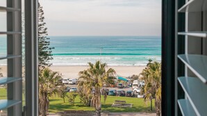 Vista de uma praia vista da janela, com cortinas, mostrando o oceano, a praia, palmeiras e carros estacionados.