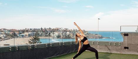 Uma pessoa fazendo uma postura de yoga em um tapete azul, com um horizonte urbano e uma praia ao fundo.
