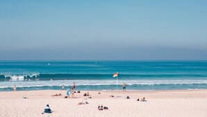 Uma praia com pessoas se banhando e tomando sol, uma bandeira na costa e um céu azul e claro.