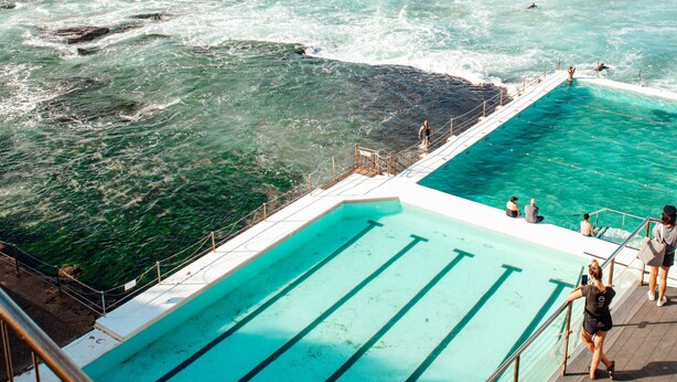 Uma piscina com água azul cristalina, uma praia e um horizonte urbano ao fundo.